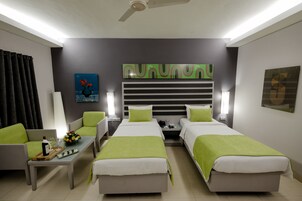 Quarto de hotel moderno com duas camas de solteiro, uma mesa de escritório, uma cadeira, uma pequena mesa com um vaso de flores e um ventilador de teto.