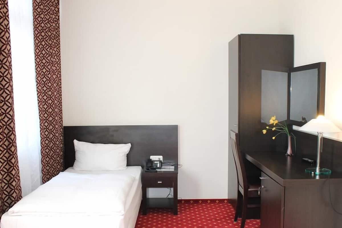 Quarto de hotel com cama, escrivaninha, cadeira, mesa de cabeceira, luminária e espelho.