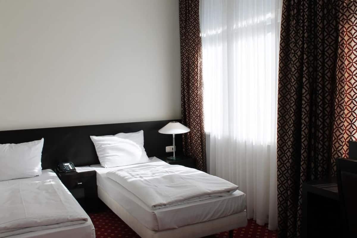 Quarto de hotel com duas camas, uma mesinha de cabeceira com abajur e cortinas estampadas.