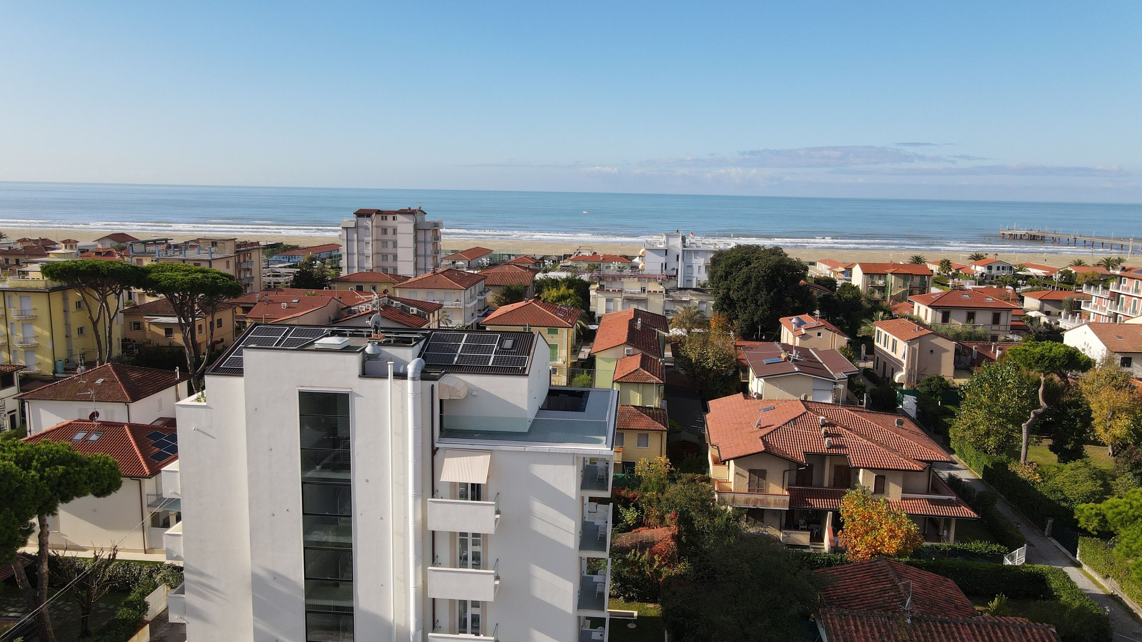 Uma área residencial costeira com edifícios de telhado vermelho, alguns com painéis solares, e vista para a praia e oceano.