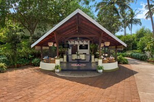 Um pavilhão coberto com uma mesa central e assentos, rodeado por vasos de plantas e um caminho de tijolos.
