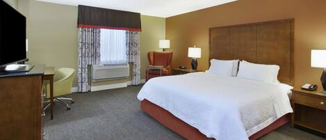 Quarto de hotel com uma cama grande, uma mesa de trabalho com televisão de tela plana, uma cadeira, duas luminárias de cabeceira e uma janela com cortinas.