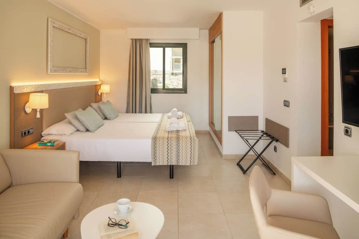 Quarto de hotel com uma cama, duas mesas de cabeceira com luminárias, uma mesinha com uma chávena e copos, uma cadeira e uma televisão.