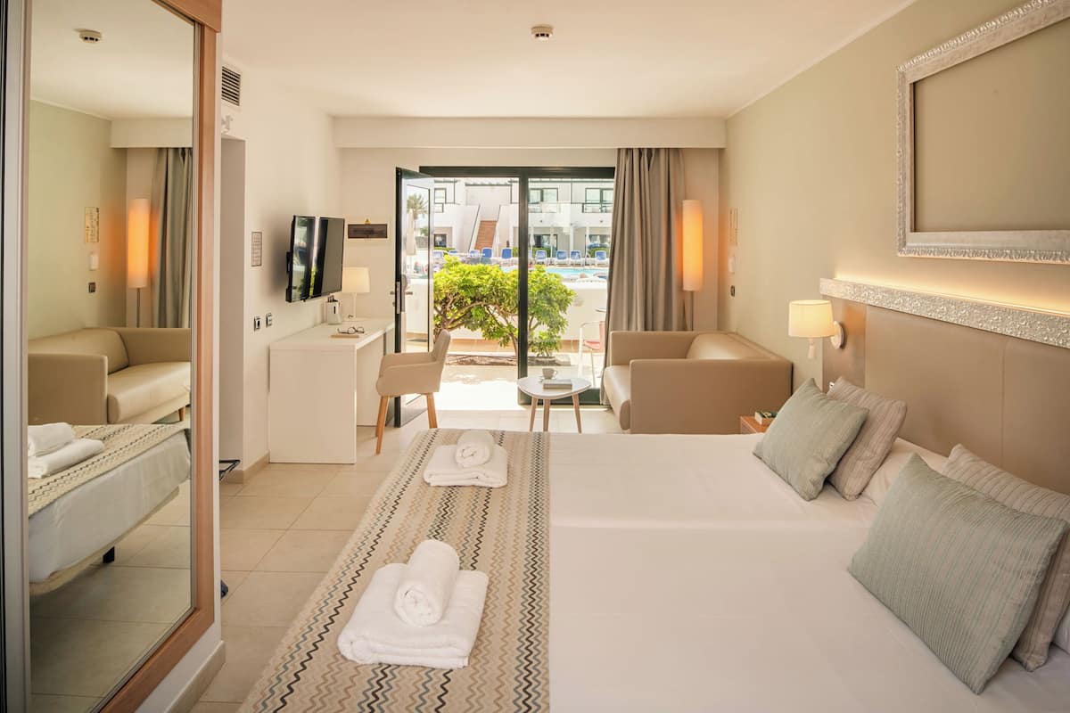 Quarto de hotel com cama, duas poltronas, uma escrivaninha, televisão e vista para a piscina.