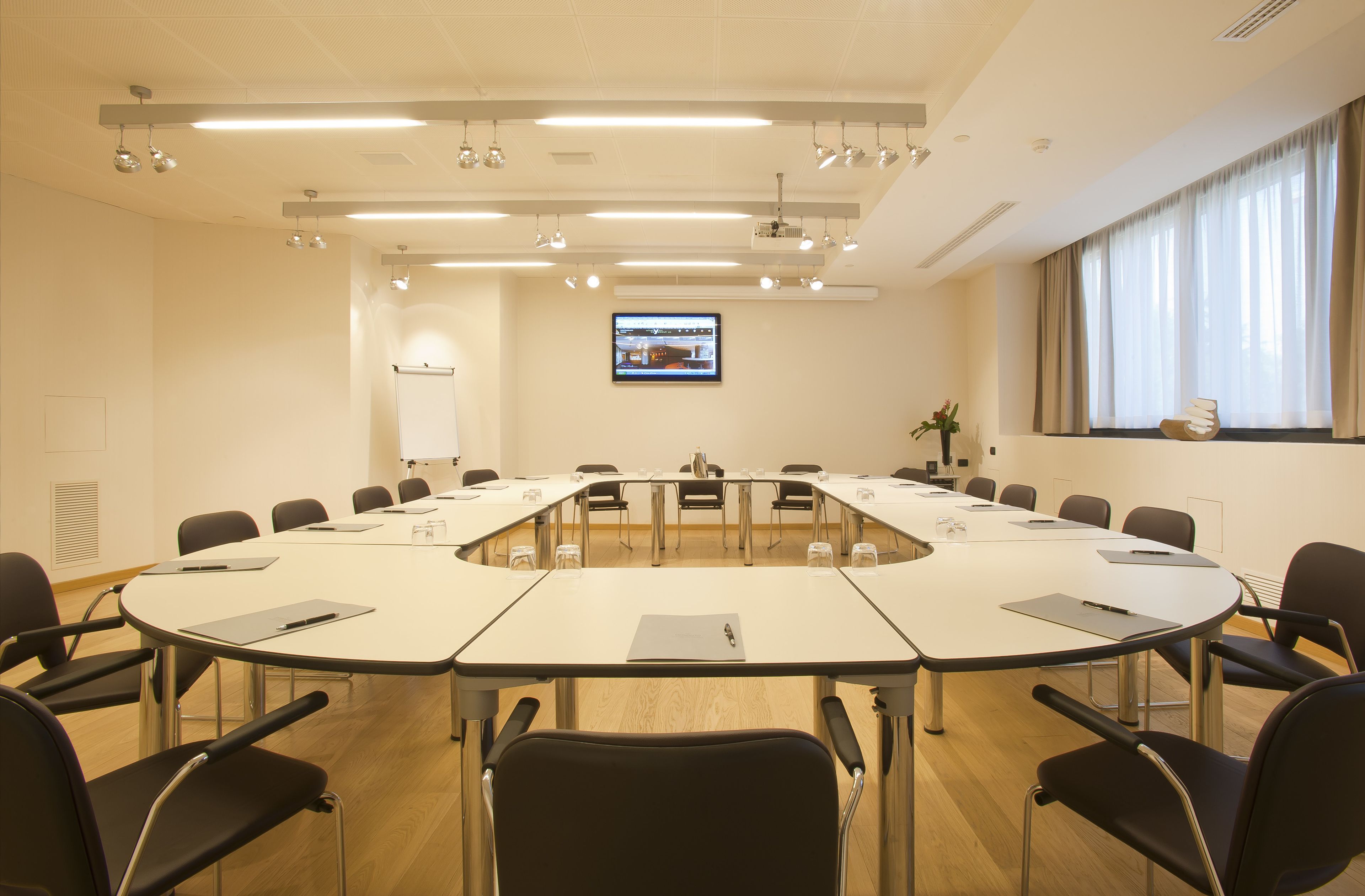 Uma sala de conferências com uma mesa comprida, cadeiras, tela de projetor e uma janela com cortinas.