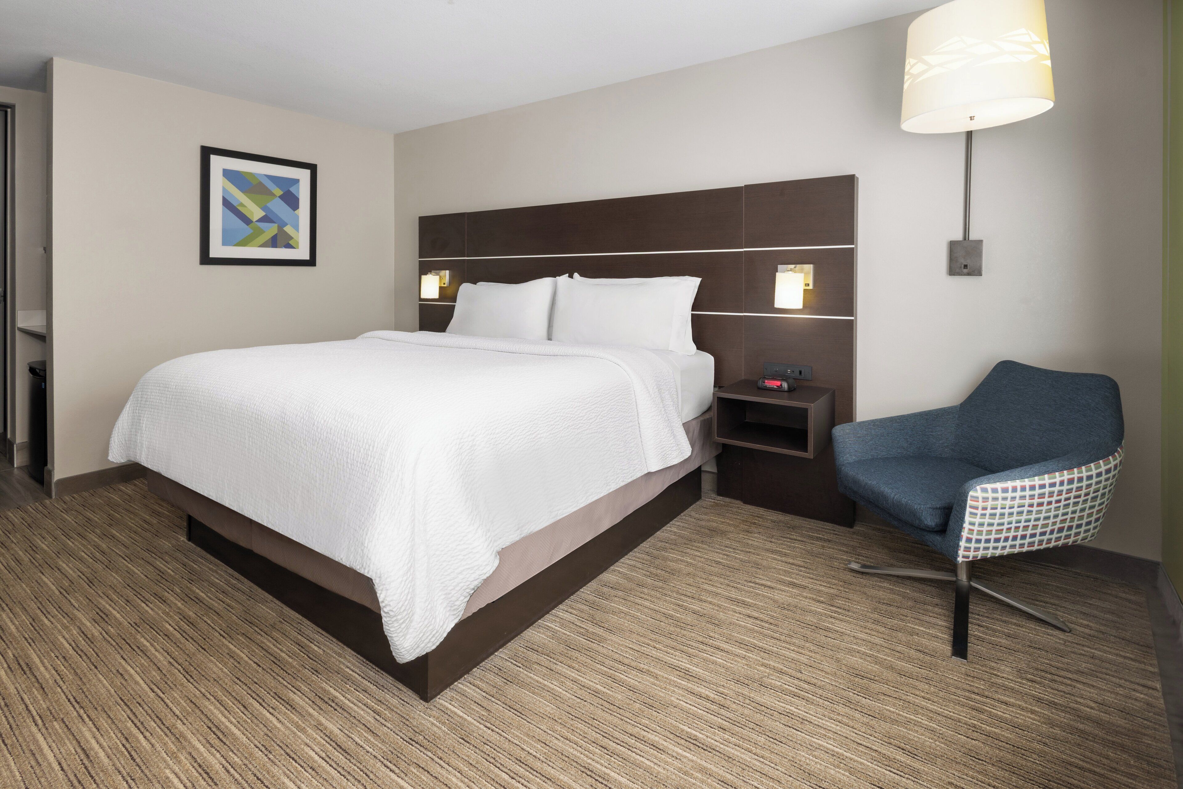Quarto de hotel com uma cama grande, uma escrivaninha com cadeira, uma luminária de parede e um quadro emoldurado.