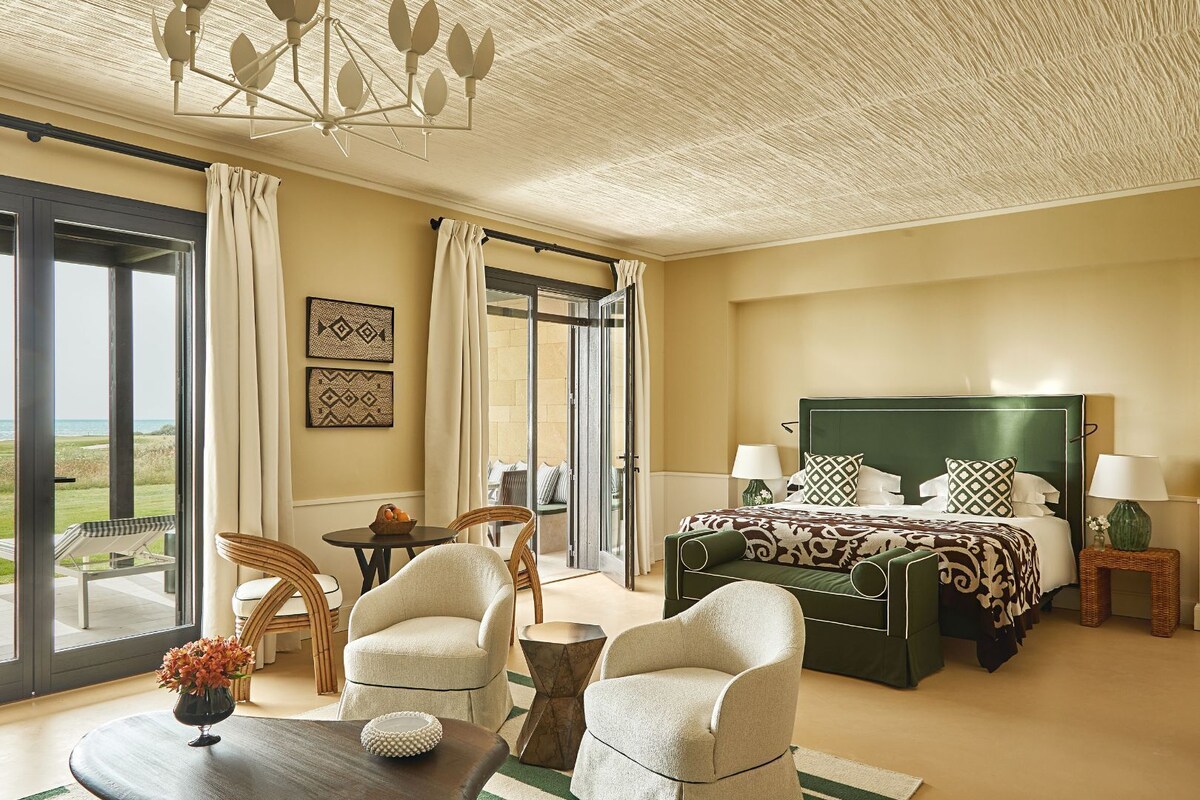 Quarto com cama, duas cadeiras, mesa, abajur e vista para o exterior.