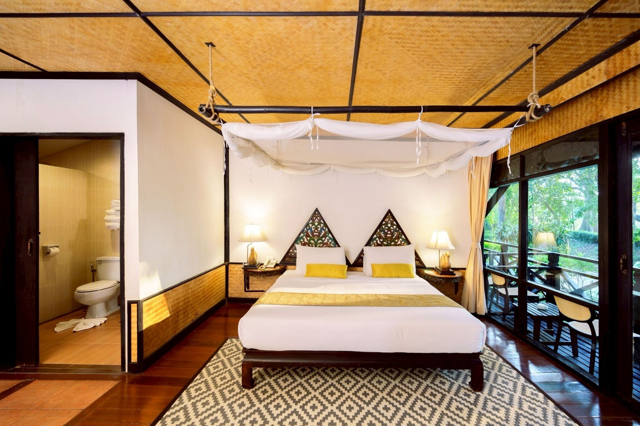 Quarto de hotel com cama grande, duas mesas de cabeceira, dossel e vista para o exterior, além de banheiro com vaso sanitário e pia.