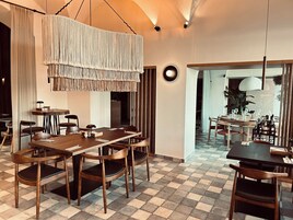 Uma sala de jantar com piso xadrez, mesas e cadeiras de madeira, e um lustre.