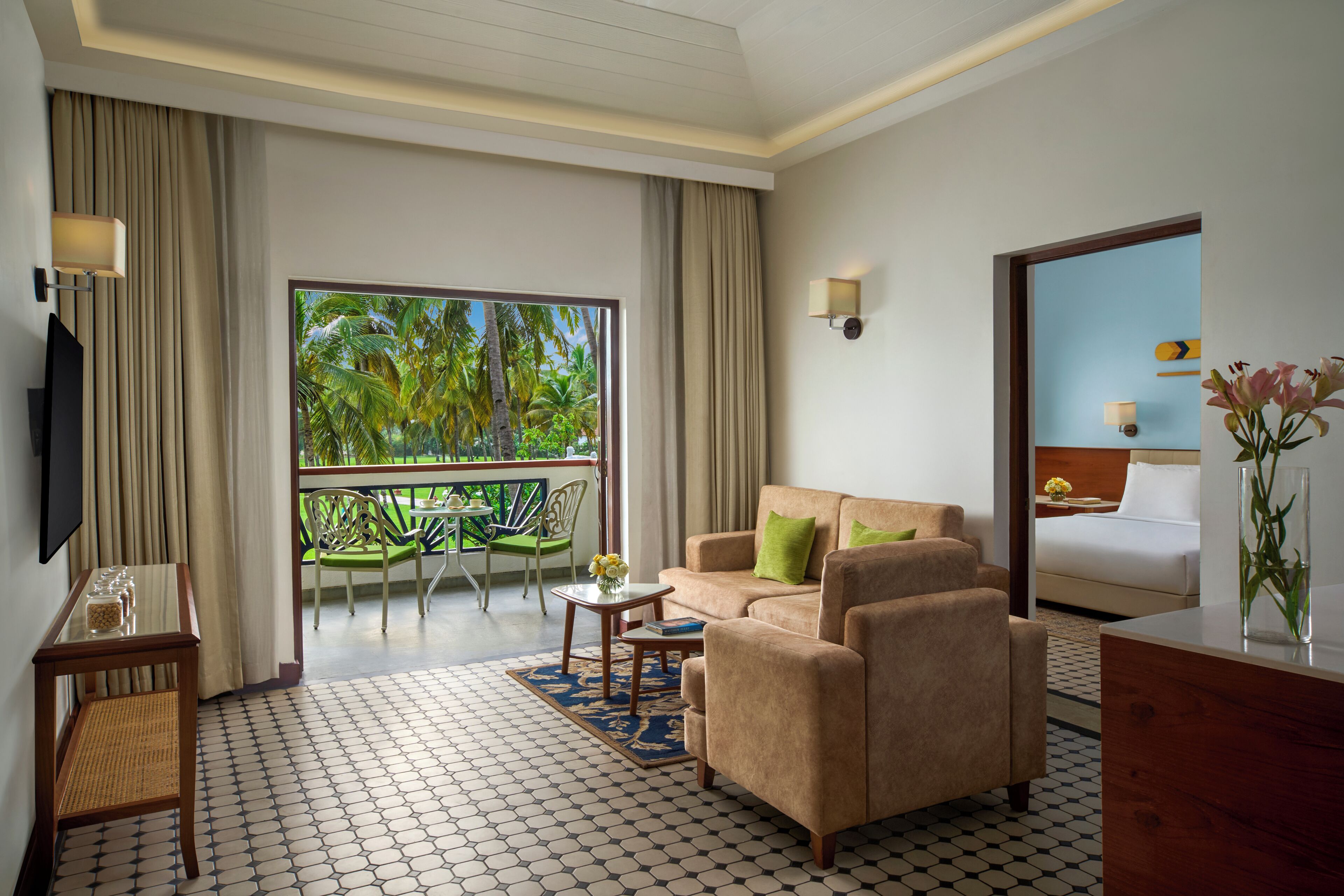 Quarto de hotel com varanda, sofá, poltrona, mesa pequena e televisão.