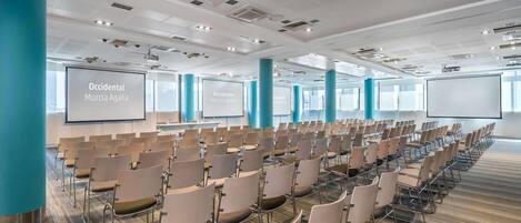 Uma sala de conferências com fileiras de cadeiras, uma tela grande e um projetor.