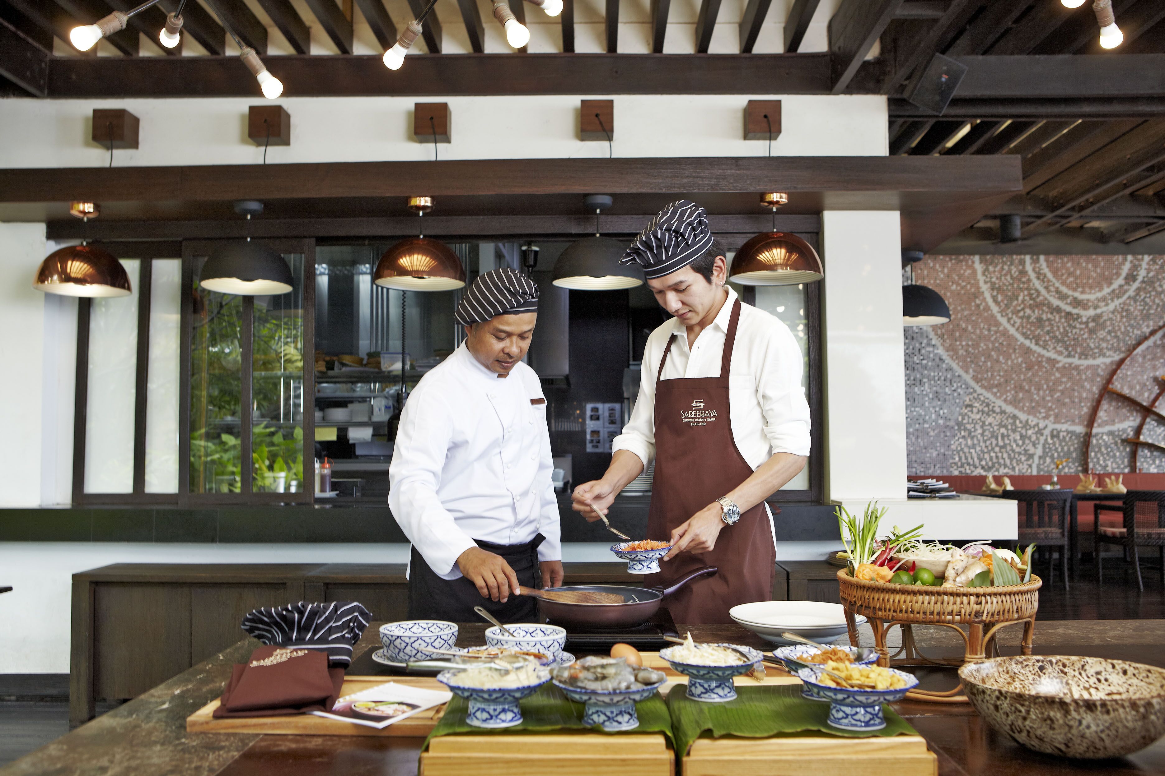 Dois chefs preparando comida em uma cozinha moderna com vigas de madeira e iluminação pendente.