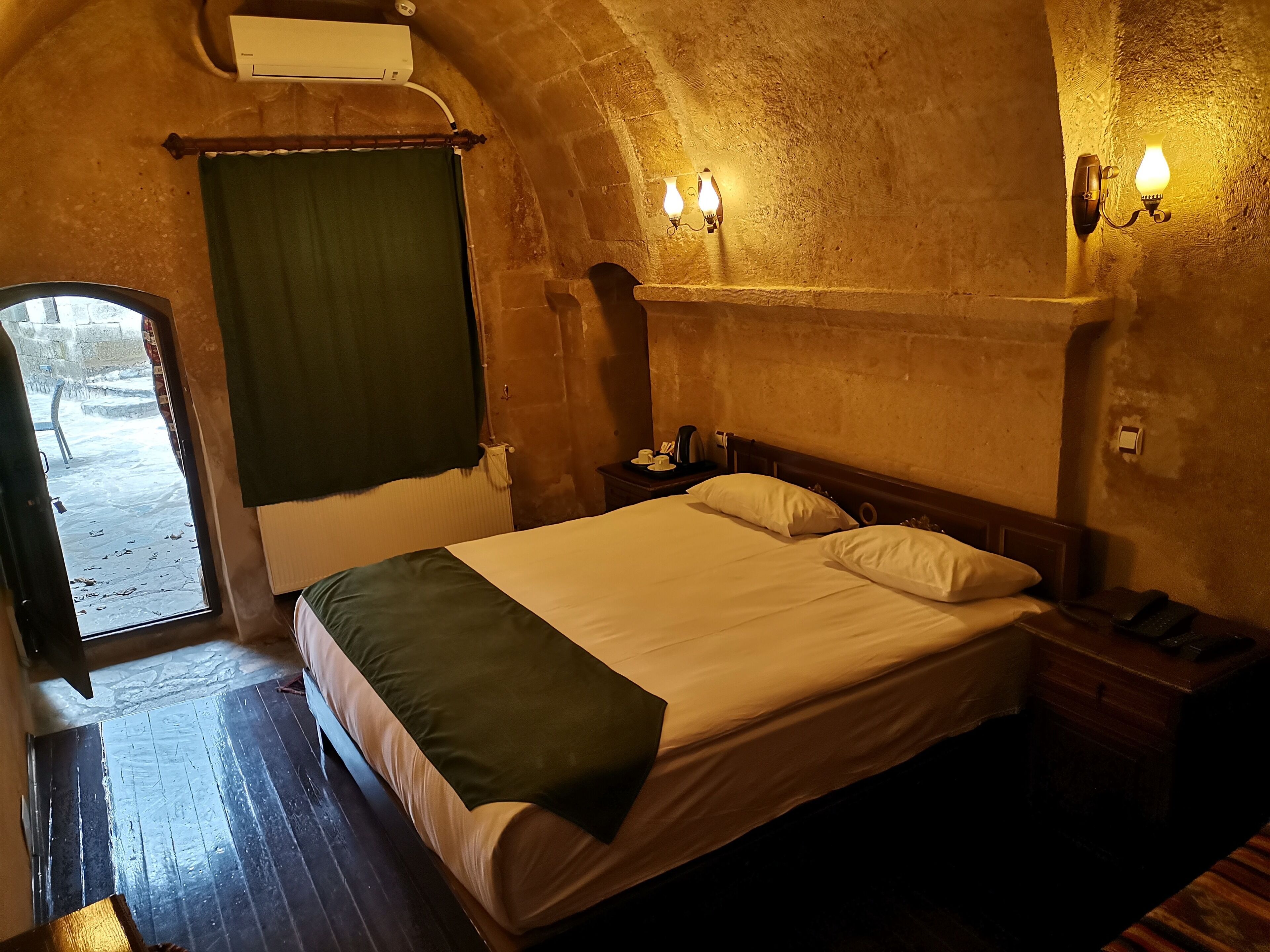 Um quarto aconchegante, com iluminação suave, possui uma cama de solteiro, uma mesinha de cabeceira e um aparelho de luz fixado na parede.