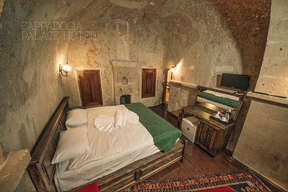 Quarto com cama, uma mesinha e televisão, e luminária fixada na parede.