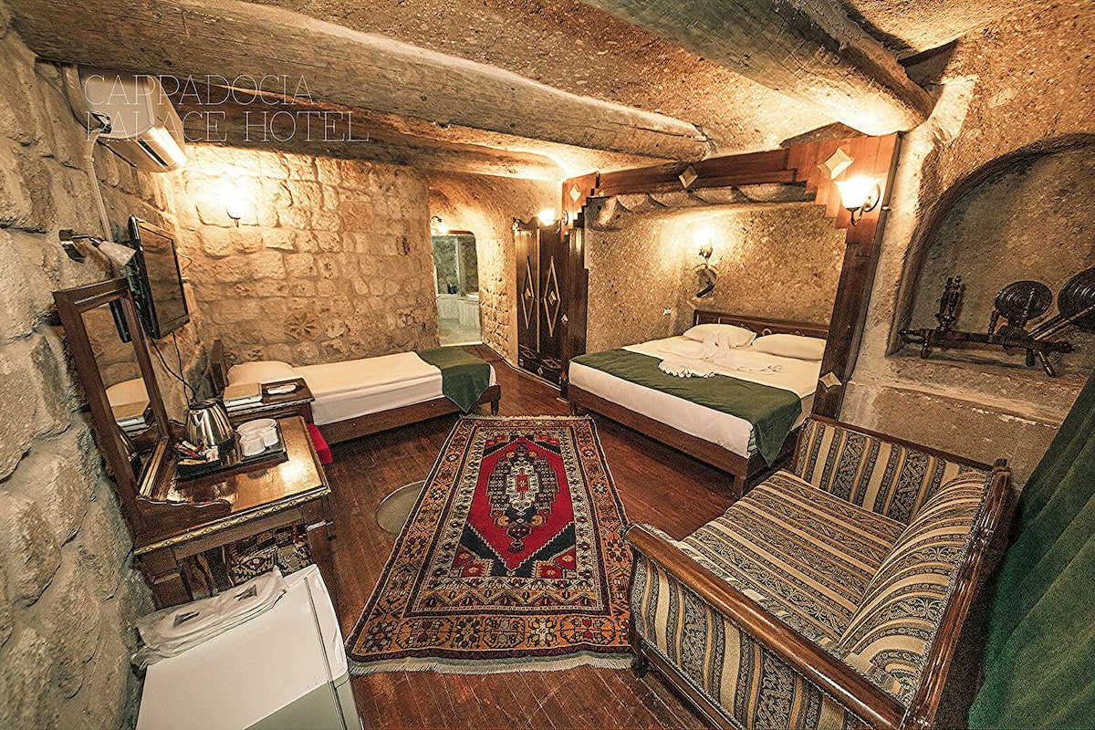 Quarto tradicional com paredes de pedra, cama de madeira, uma mesinha com chaleira e tapete estampado.