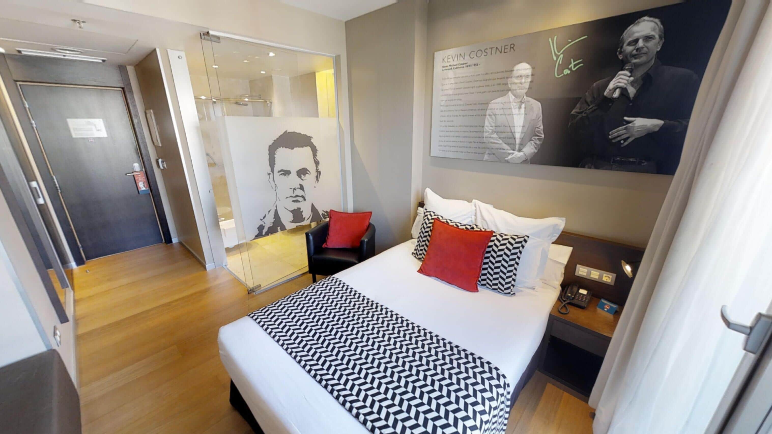 Quarto de hotel com cama, escrivaninha, cadeira, divisória de vidro e um cartaz na parede.