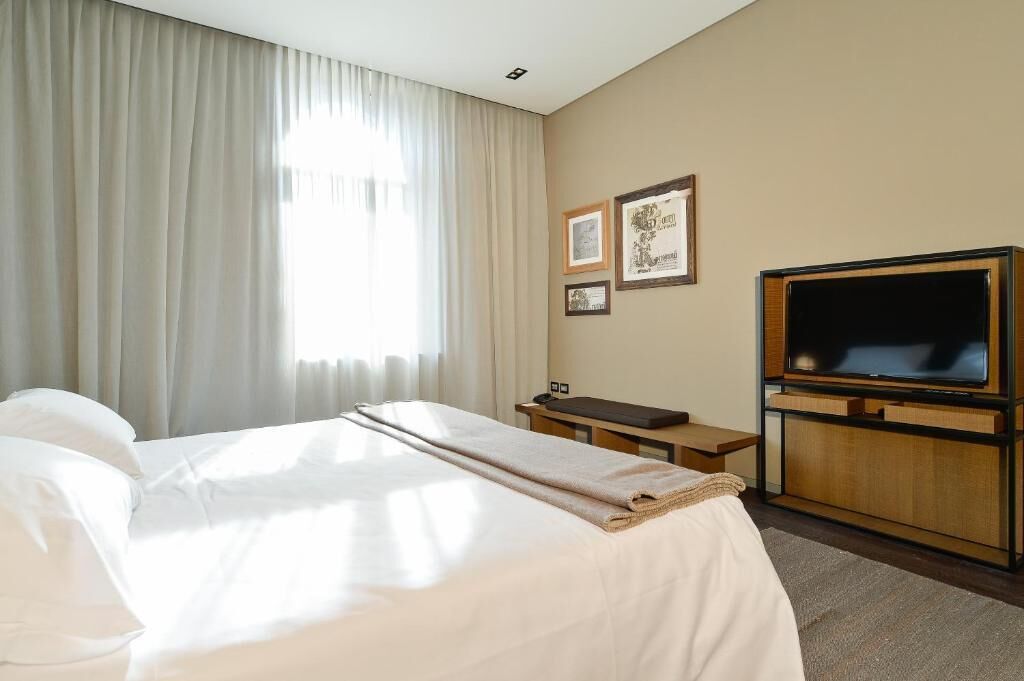 Quarto de hotel com cama, televisão, escrivaninha e janela com cortinas.