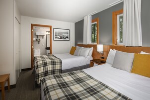 Quarto de hotel com duas camas, uma mesinha, uma mesa de cabeceira, uma janela com cortinas, um quadro na parede e um banheiro visível por uma porta aberta.