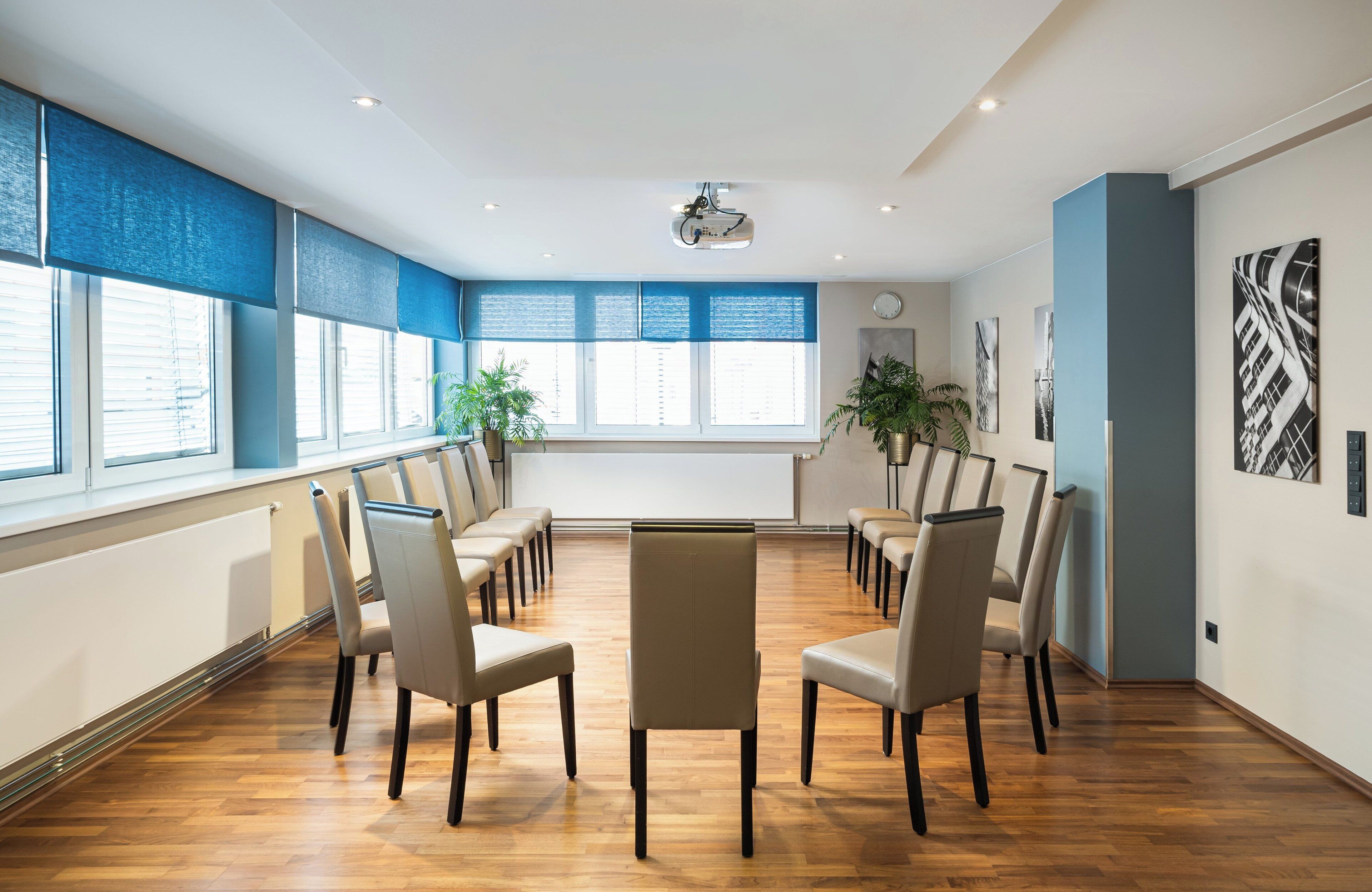 Sala de conferências com piso de madeira, cadeiras bege, projetor e janelas amplas com persianas.