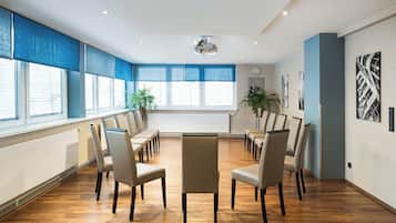 Sala de conferências com piso de madeira, cadeiras bege, projetor e janelas amplas com persianas.