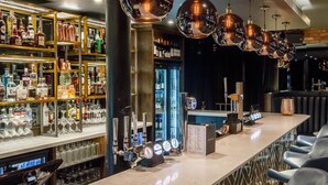 Uma área de bar com prateleiras cheias de garrafas de bebidas, um balcão com diversas torneiras e uma área de estar com cadeiras.