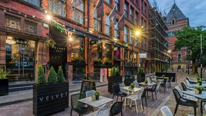 Área externa com mesas e cadeiras, uma placa com o nome 'VELVET HOTEL' e fachada de um edifício de tijolos com iluminação decorativa.