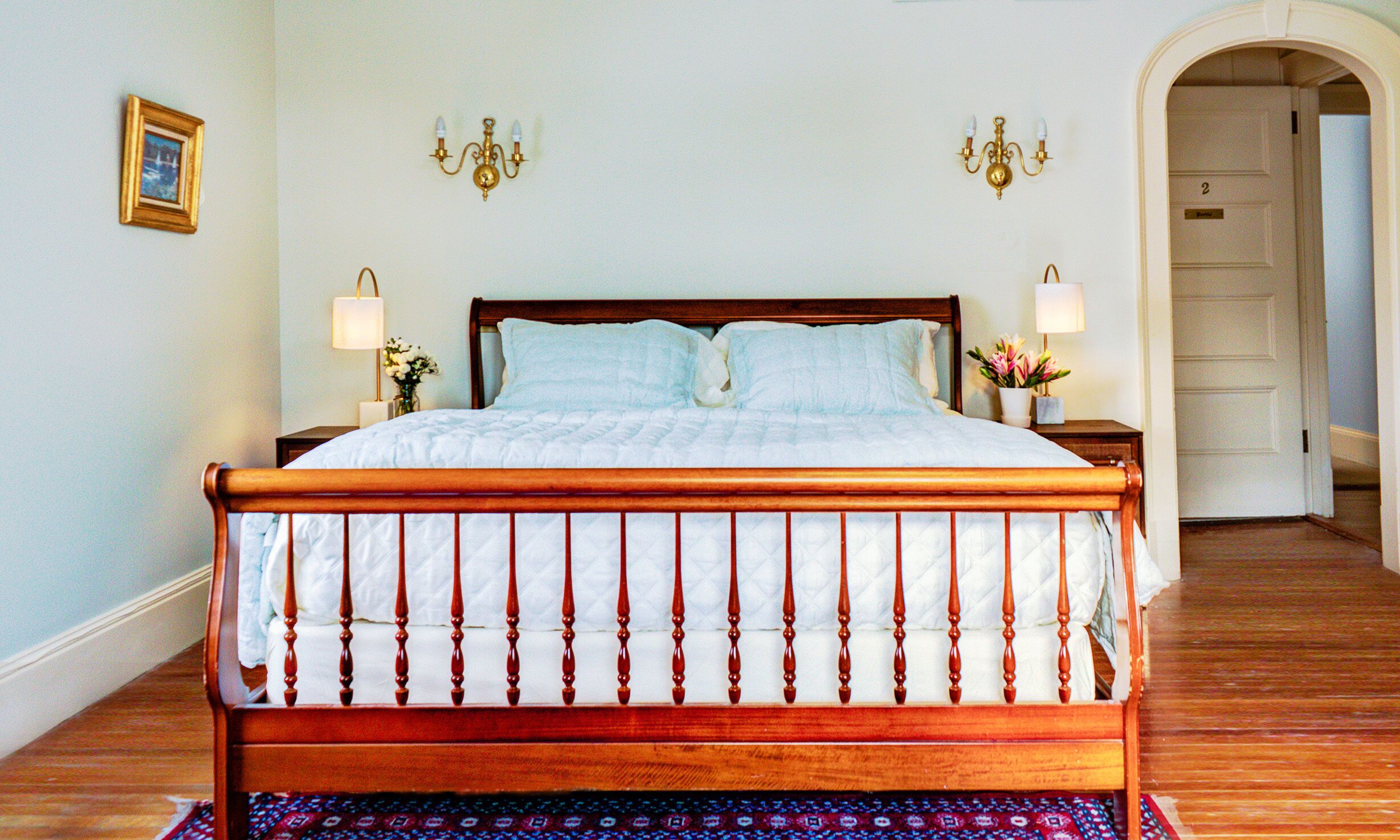 Uma cama de madeira grande com edredão branco acolchoado, duas mesas de cabeceira com abajures e um quadro na parede.