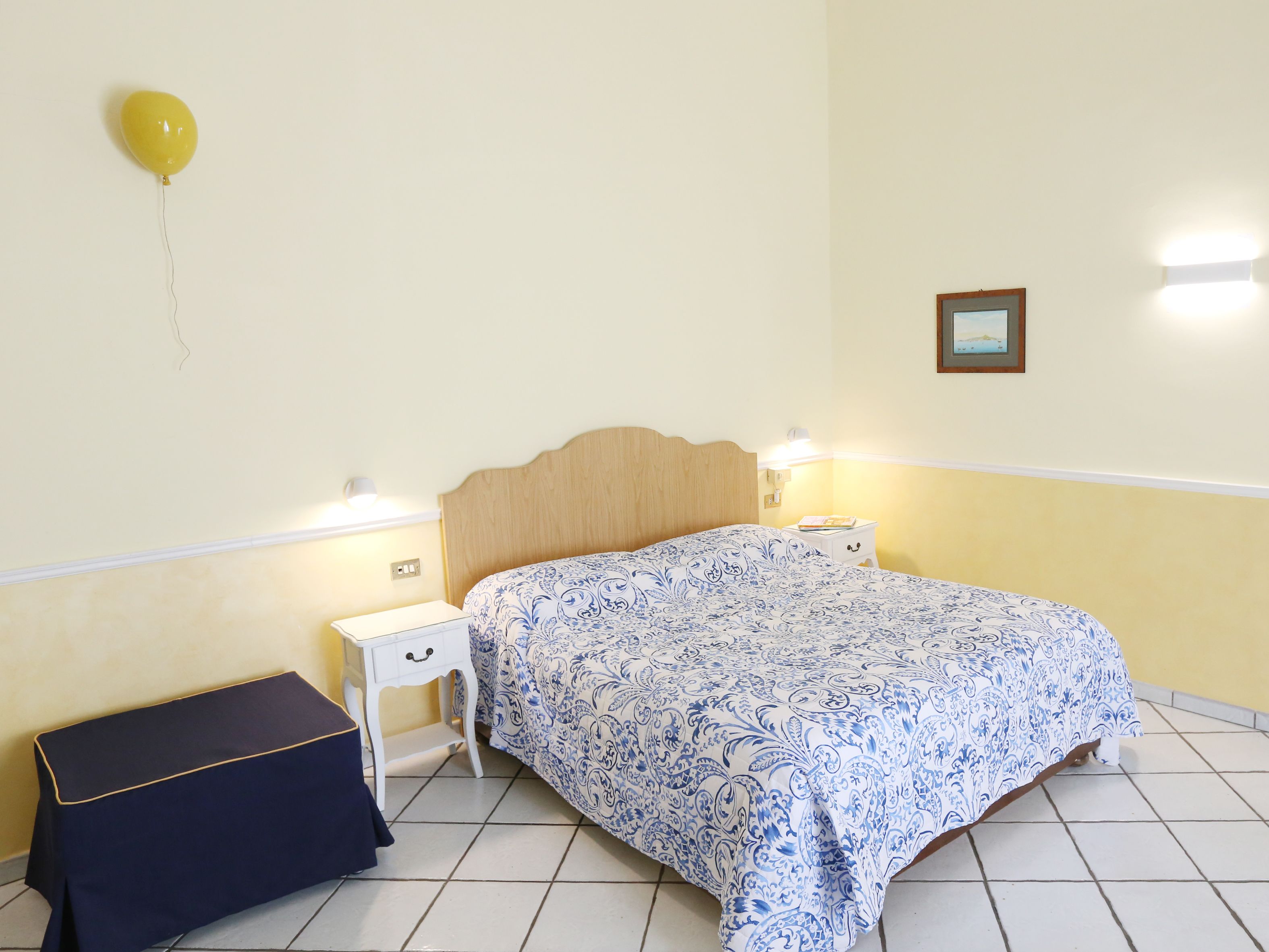 Quarto com cama, mesas de cabeceira, um criado-mudo, uma mesinha e um quadro na parede, além de um balão.