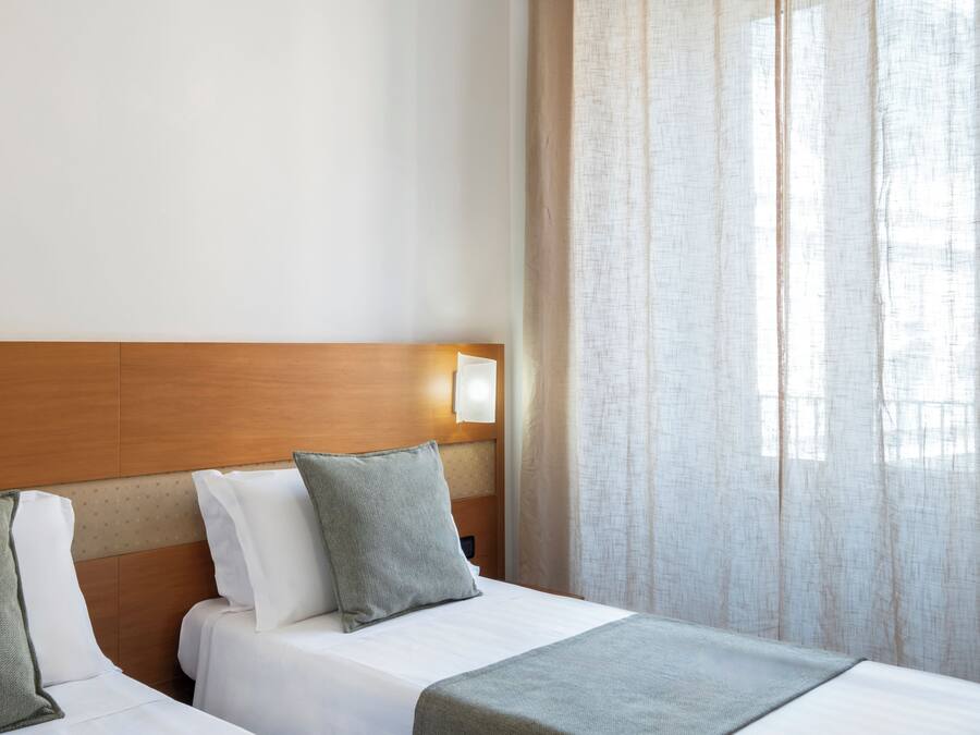 Quarto de hotel com cabeceira de madeira, duas camas, mesa de cabeceira, abajur e cortinas leves.