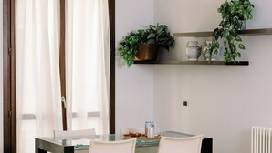 Uma área de refeições com mesa de vidro, cadeiras brancas, um sofá verde e um espaço de cozinha com radiador e prateleiras com plantas.
