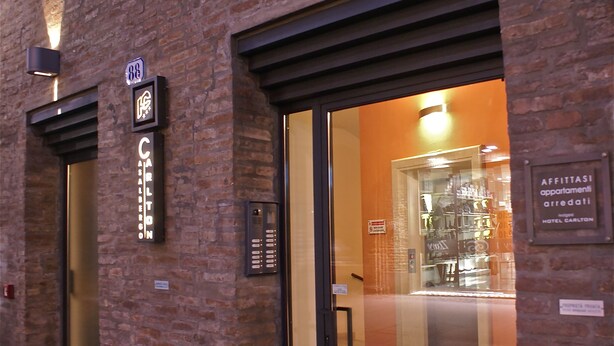 Entrada de hotel com fachada de tijolos, um elevador e uma porta de vidro que dá acesso ao interior.