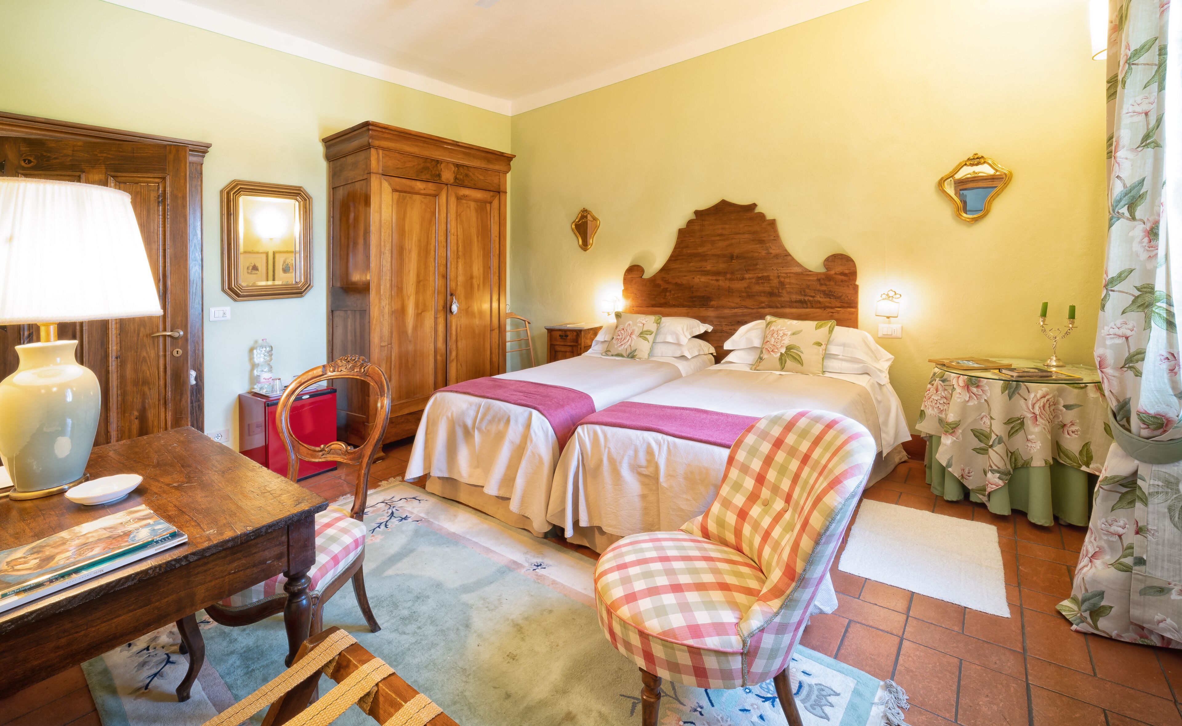 Cama de casal com roupa de cama florida, guarda-roupa de madeira, escrivaninha com abajur e cadeira com almofada xadrez.
