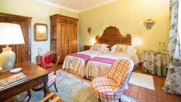 Cama de casal com roupa de cama florida, guarda-roupa de madeira, escrivaninha com abajur e cadeira com almofada xadrez.