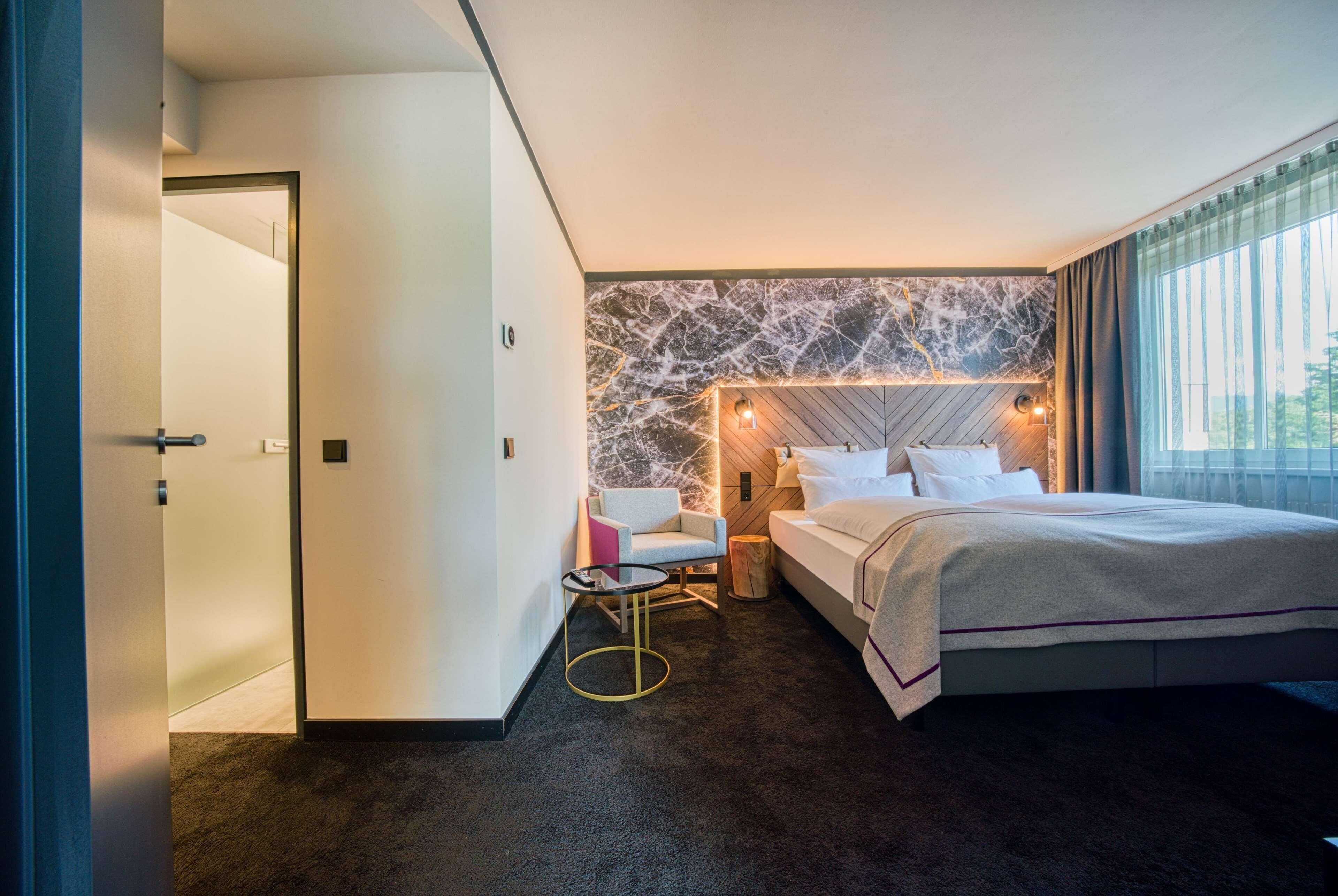 Quarto de hotel com uma cama grande, área de estar com uma cadeira e mesa, parede decorativa em mármore e vista para o exterior.