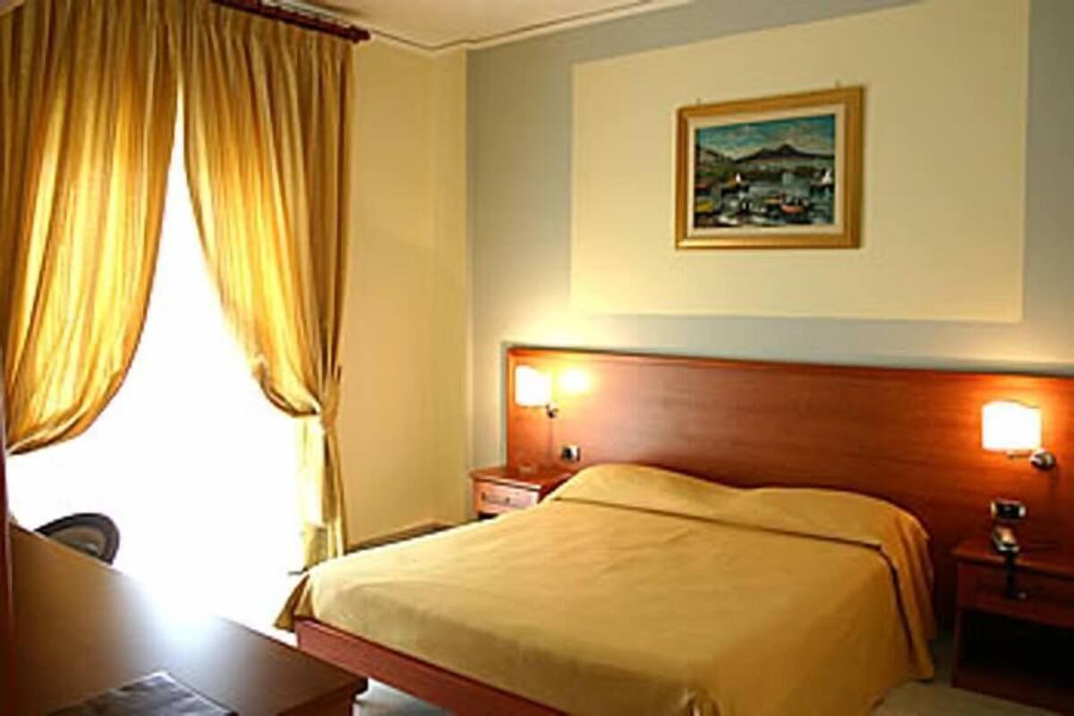 Quarto de hotel com cama, mesa de cabeceira, luminária, um quadro na parede e janela com cortinas.