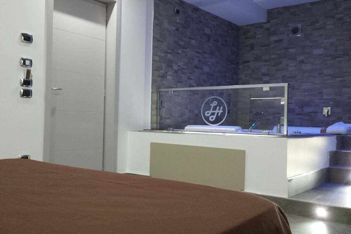Quarto de hotel moderno com cama, banheiro com box de vidro e parede texturizada.