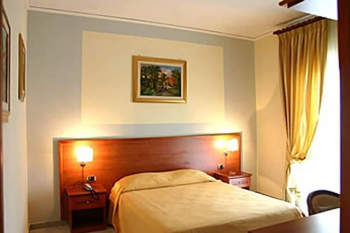 Quarto de hotel com cabeceira de madeira, duas luminárias de cabeceira, cama com manta bege, mesa de cabeceira com telefone e um quadro na parede.