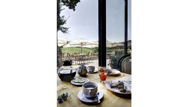 Mesa posta para o café da manhã, com pratos, xícaras e um jarro de suco, com vista para um jardim com guarda-sóis.