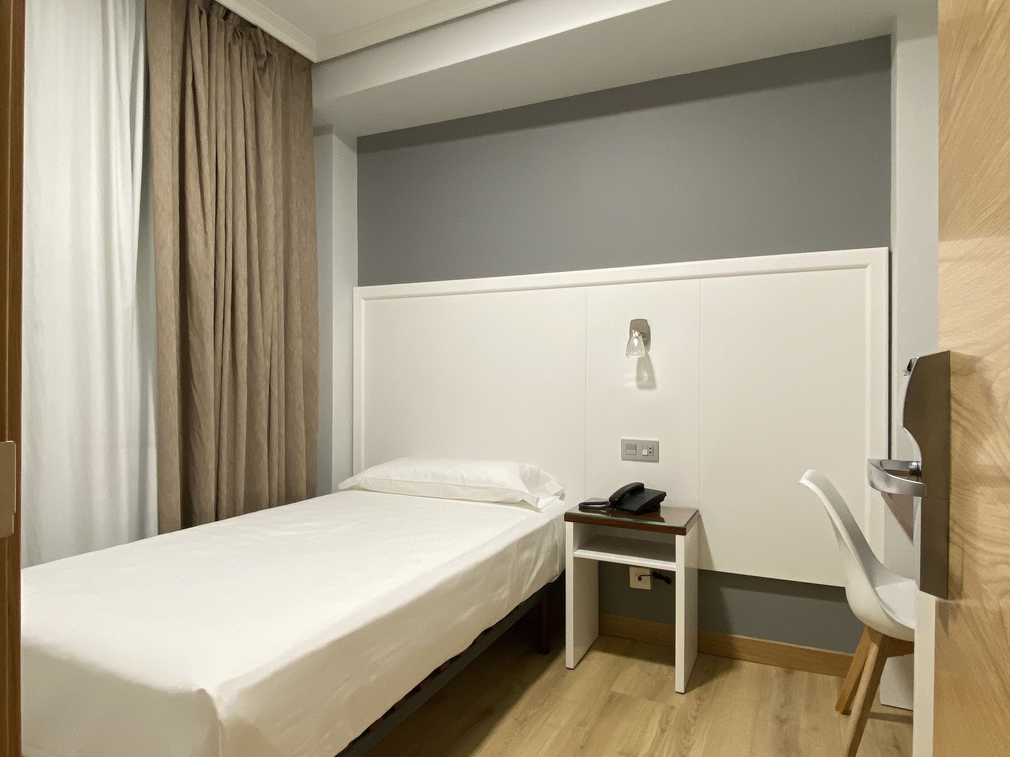 Uma cama de solteiro com colchão e almofadas brancas, uma mesinha de cabeceira com telefone e uma cadeira.