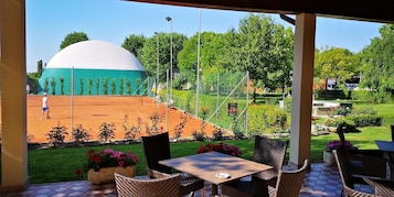 Área externa com assentos de vime e uma mesa, com vista para uma quadra de tênis e uma grande estrutura em cúpula ao fundo.