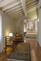 Um quarto com piso de madeira, um tapete estampado, uma cadeira, uma mesa e um abajur.