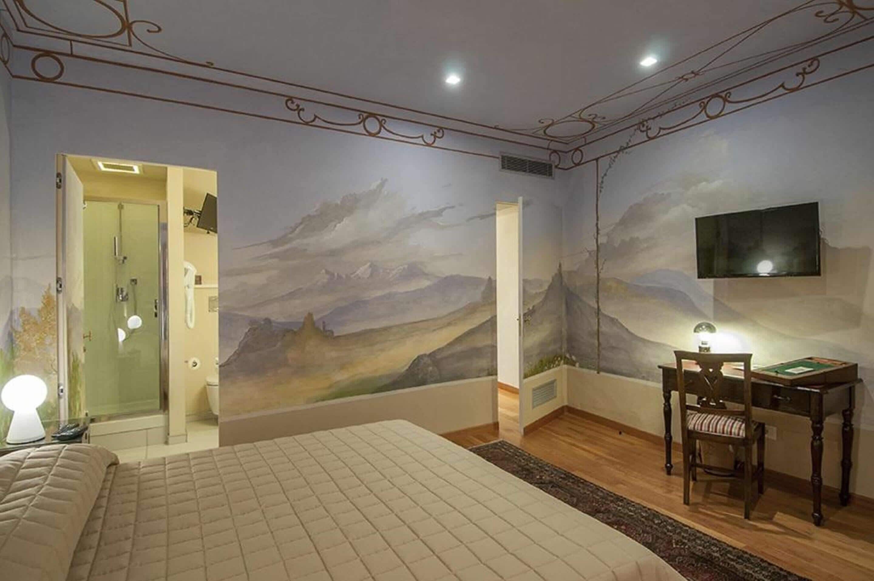 Quarto com cama, escrivaninha, cadeira, televisão e um mural na parede.