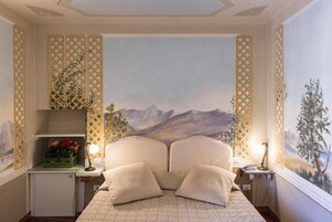 Quarto com uma cama grande, duas mesas de cabeceira, um criado-mudo, prateleira na parede com flores e um mural de montanhas.