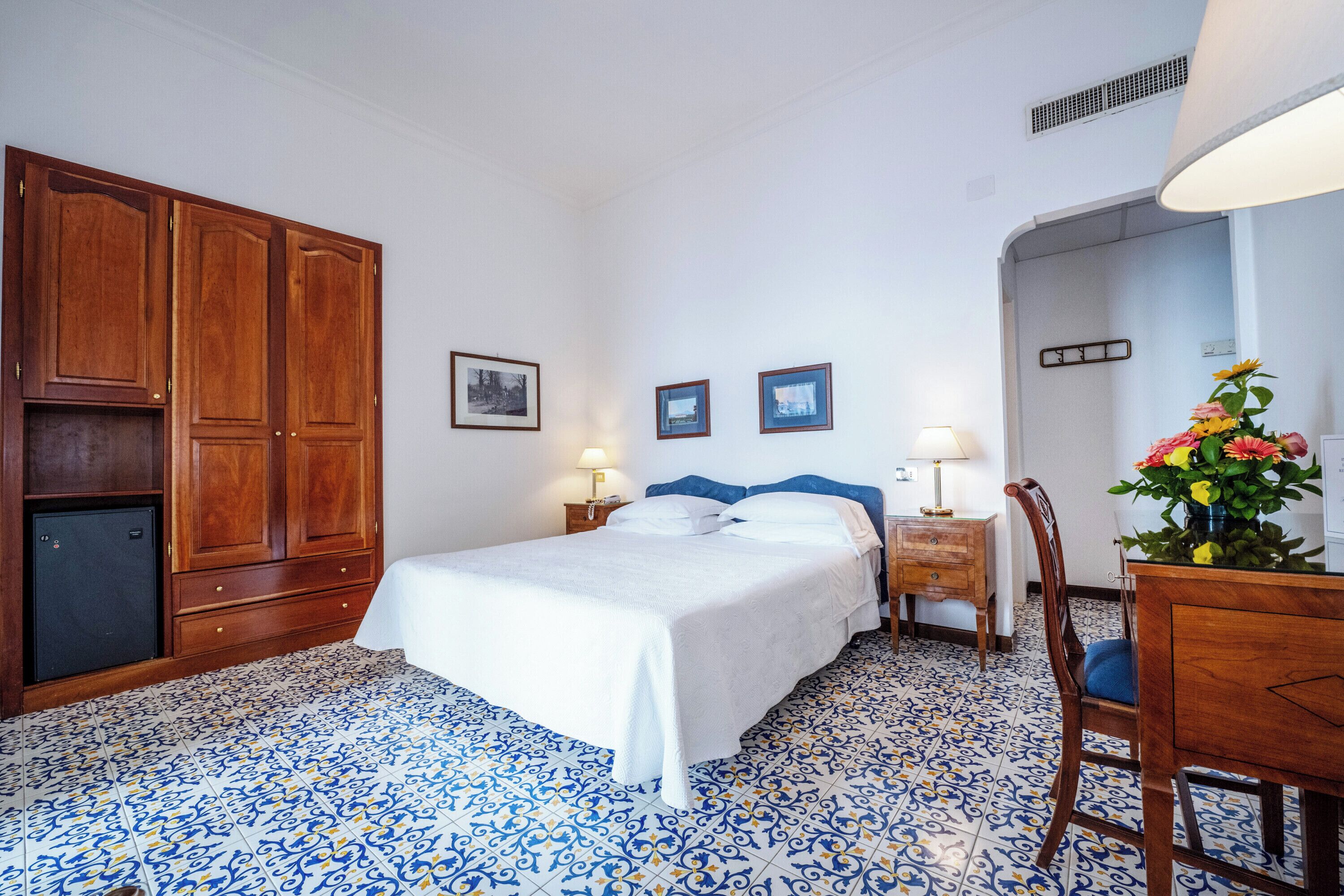 Quarto de hotel com cama, guarda-roupa de madeira, duas mesas de cabeceira com abajures, escrivaninha com cadeira e um vaso de flores.