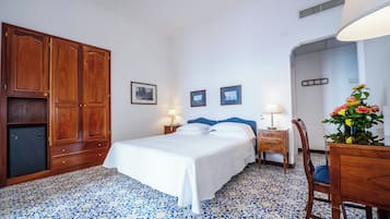 Quarto de hotel com cama, guarda-roupa de madeira, duas mesas de cabeceira com abajures, escrivaninha com cadeira e um vaso de flores.
