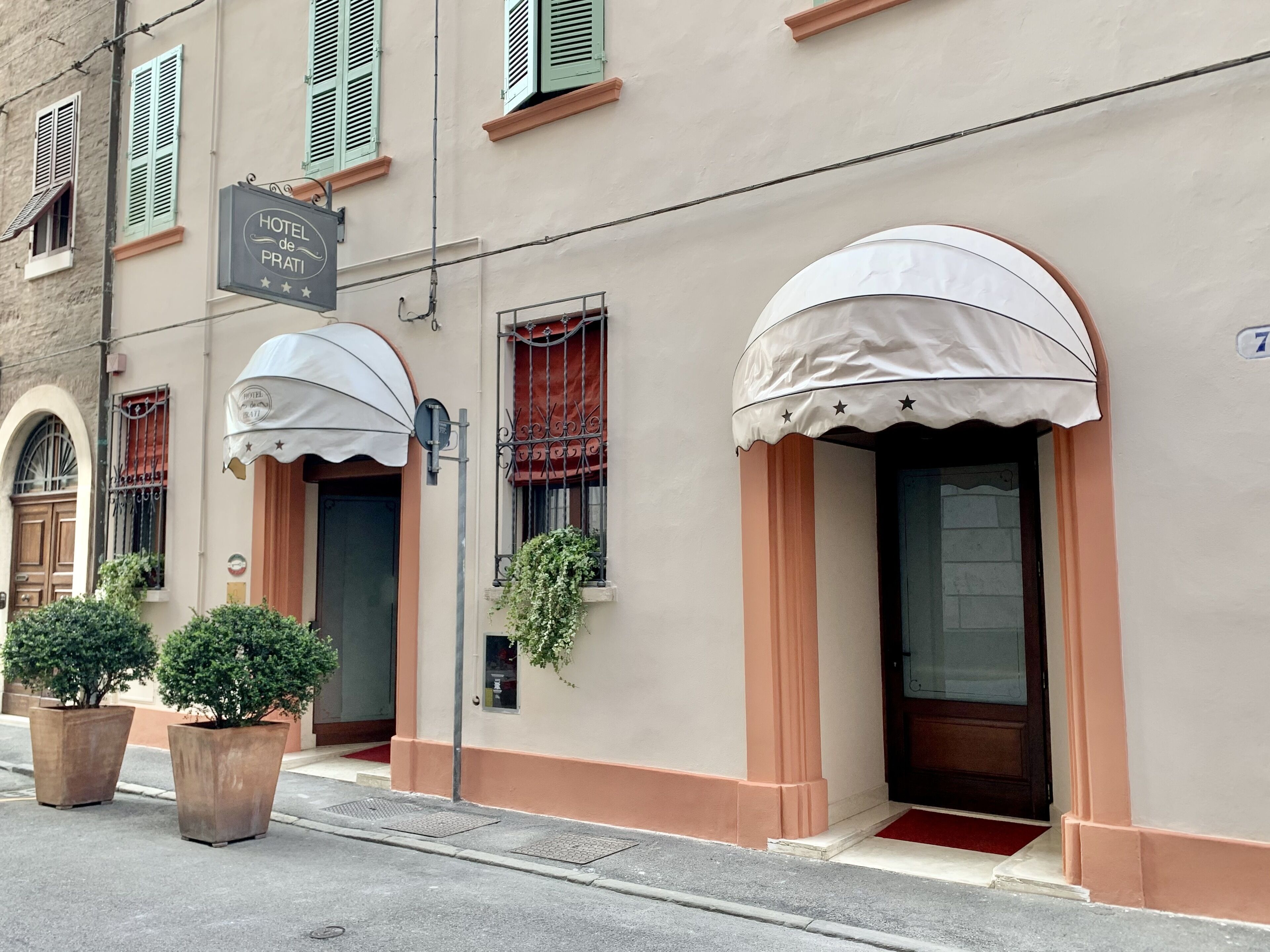 Fachada de hotel com portas em arco, uma placa com a inscrição "HOTEL de PRATI" e plantas em vasos na frente.