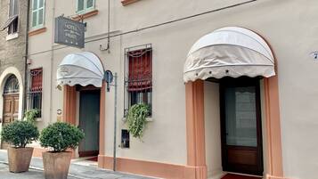Fachada de hotel com portas em arco, uma placa com a inscrição "HOTEL de PRATI" e plantas em vasos na frente.