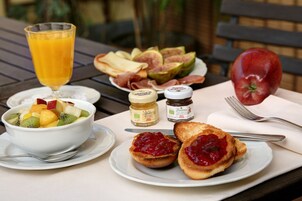 Uma mesa de café da manhã com uma tigela de frutas, torradas com geleia, um copo de suco de laranja e um prato com carnes e figos.