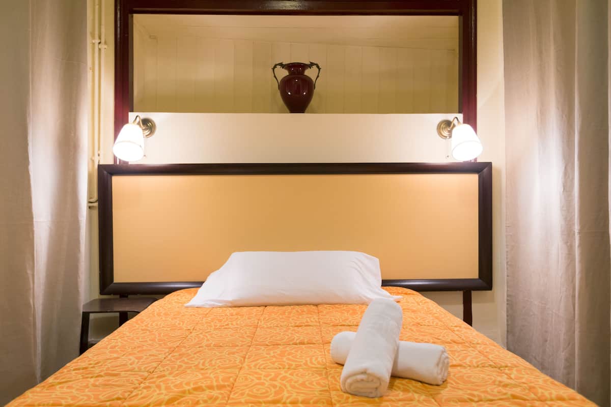 Quarto de hotel com cama, travesseiro branco, toalha enrolada e duas luminárias fixadas na parede.