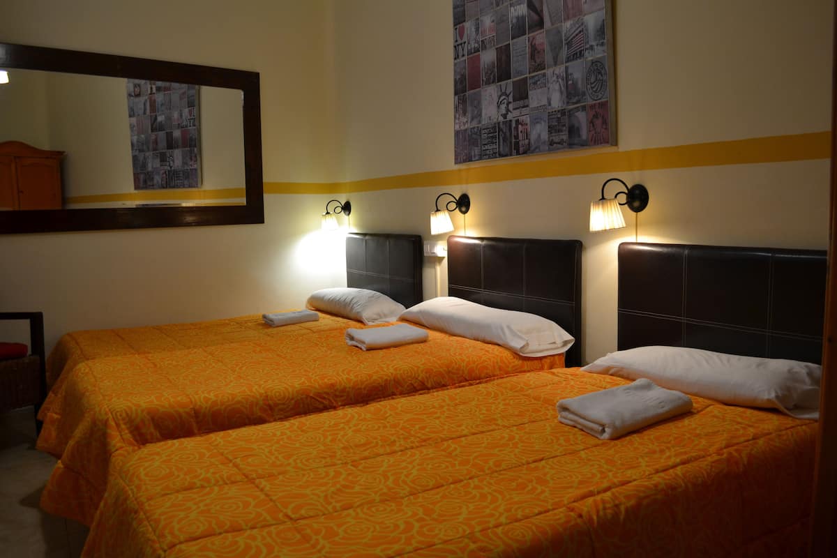 Quarto de hotel com duas camas, cada uma com colcha laranja estampada, almofadas brancas e toalhas dobradas. Há luminárias fixadas na parede acima de cada cama, e um espelho grande na parede à esquerda.